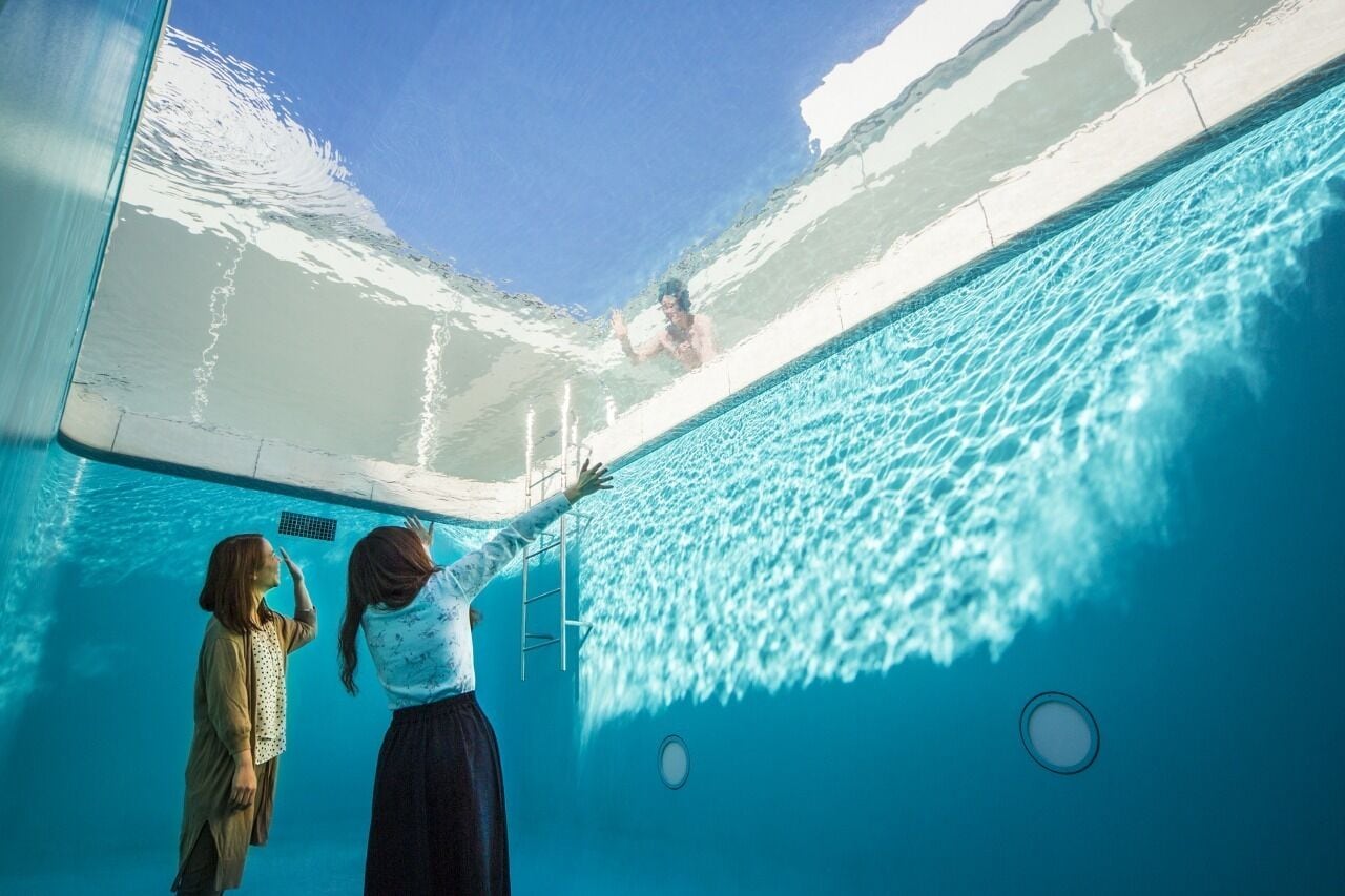 Uma piscina interior com água cristalina, uma escada e uma pessoa a nadar.