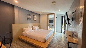Quarto de hotel moderno com uma cama grande, mesa de cabeceira de madeira, televisão na parede e vista para o corredor.