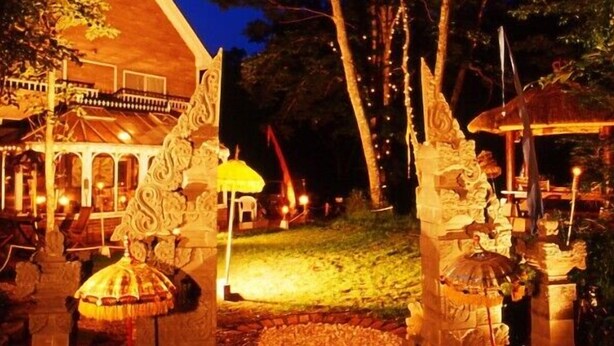 Área externa bem iluminada com estátuas de pedra, um caminho e um edifício ao fundo.