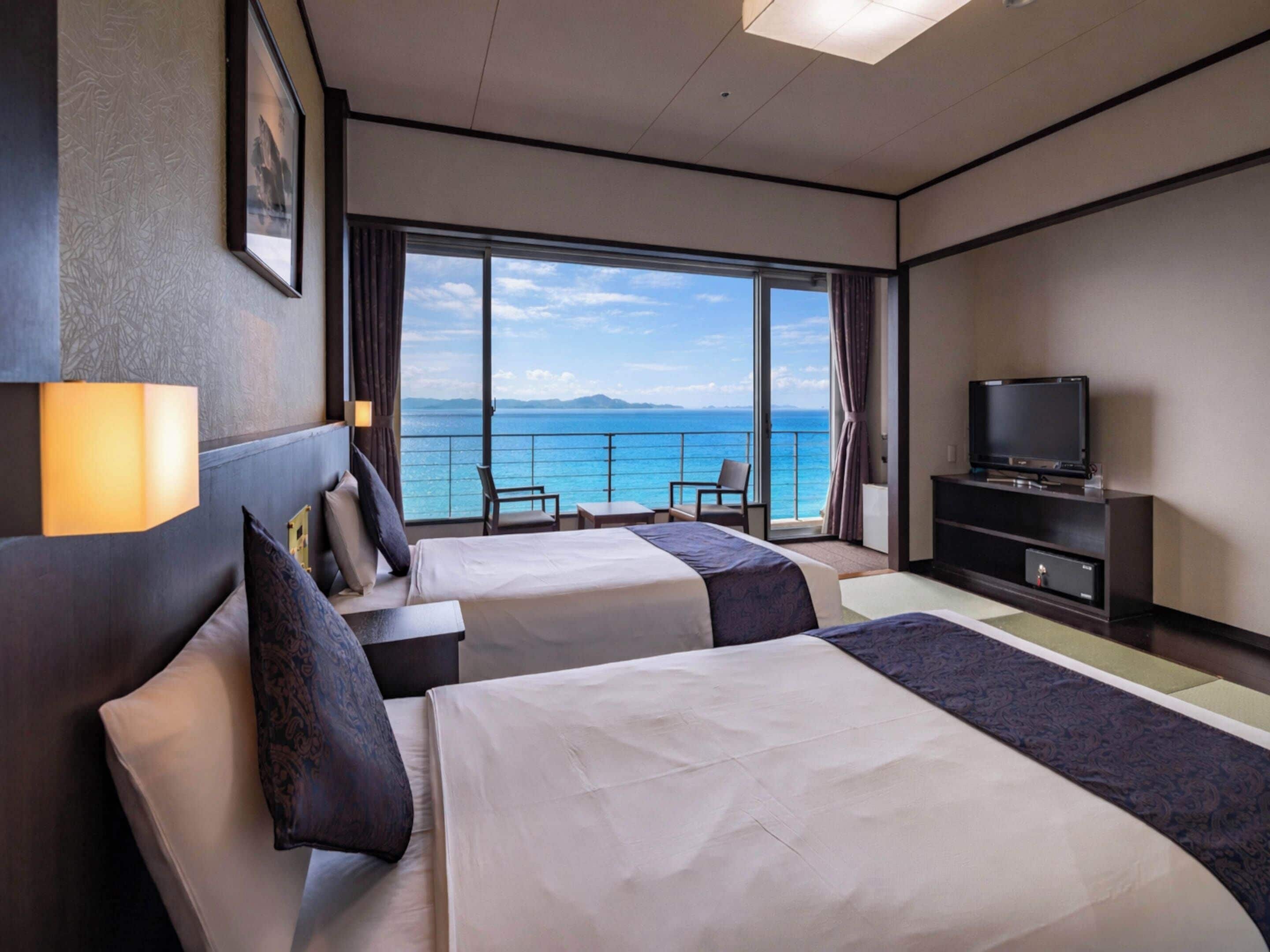 Quarto de hotel com duas camas, televisão, escrivaninha e vista para o mar.