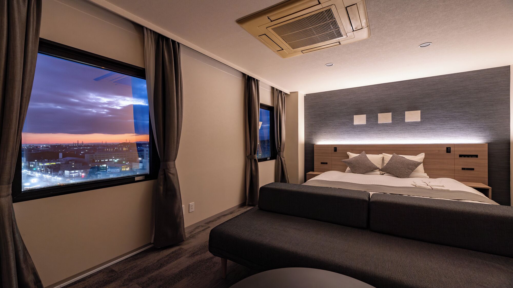 Quarto de hotel com uma janela grande, uma cama com cabeceira, um banco e uma TV fixada na parede.