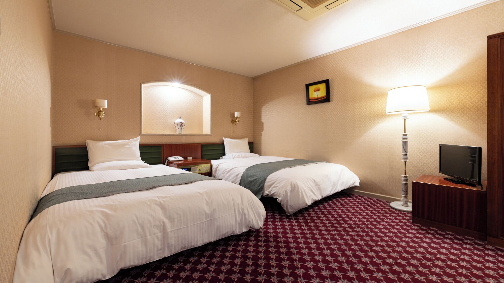 Quarto de hotel com duas camas, mesa de cabeceira, luminária, televisão e espelho.
