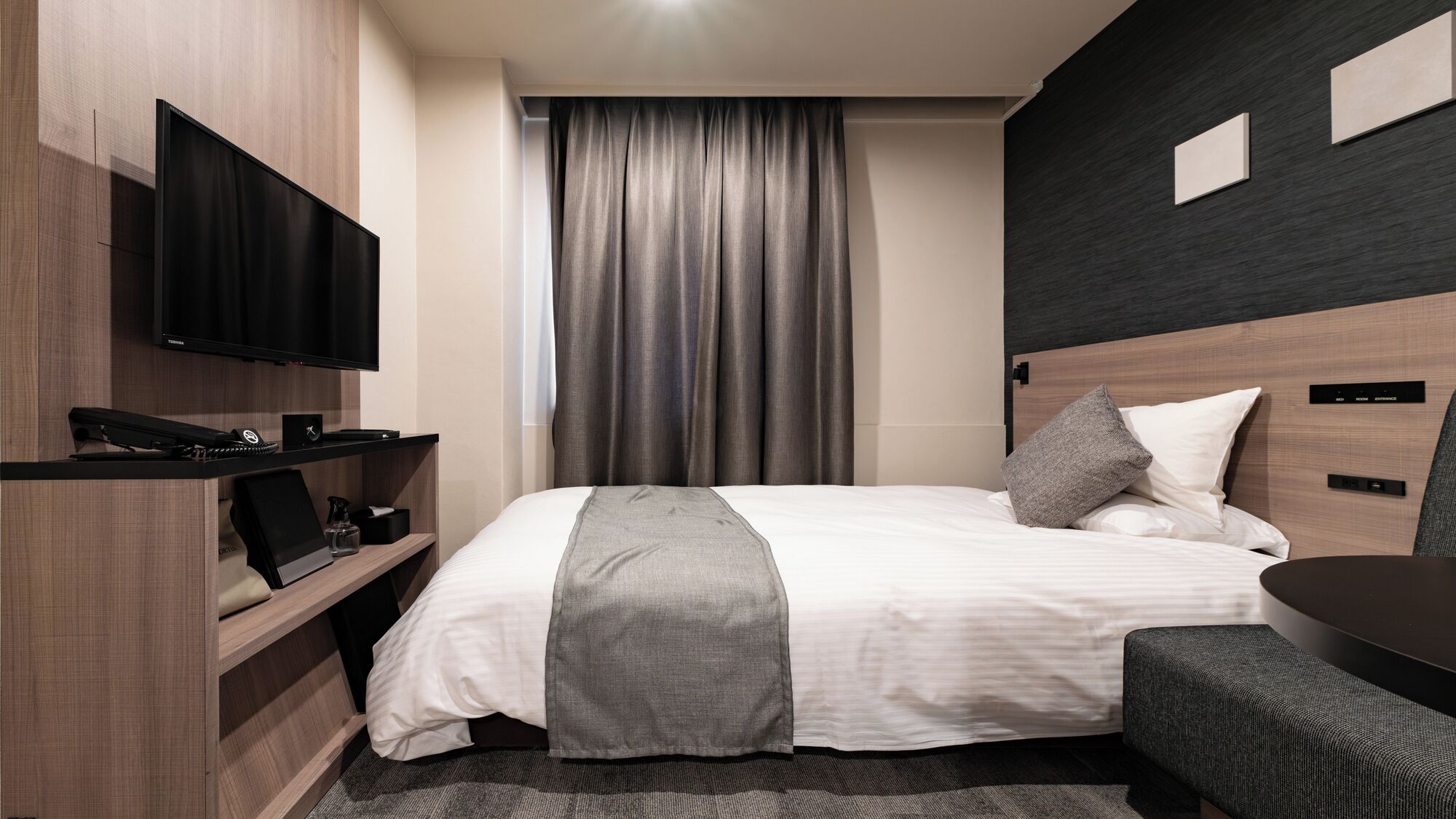 Quarto de hotel moderno com cama, escrivaninha com televisão de tela plana, cadeira e painel de controle na parede.