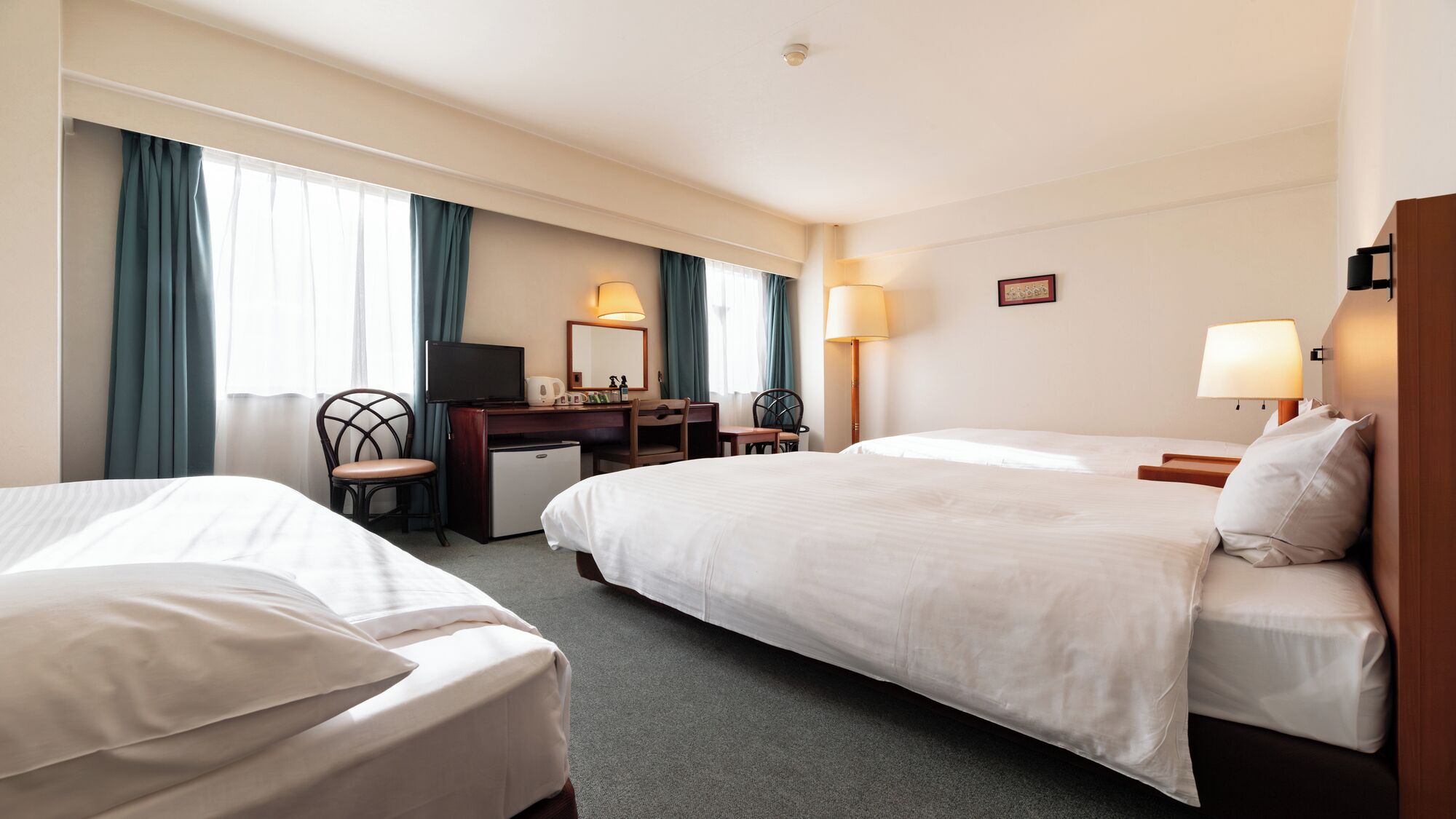 Quarto de hotel com duas camas, uma mesa com computador, uma cadeira e duas luminárias.