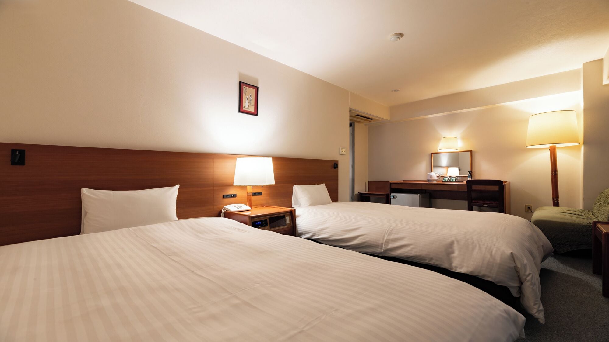 Quarto de hotel com duas camas, uma escrivaninha, uma cadeira, um abajur e uma mesinha com abajur.