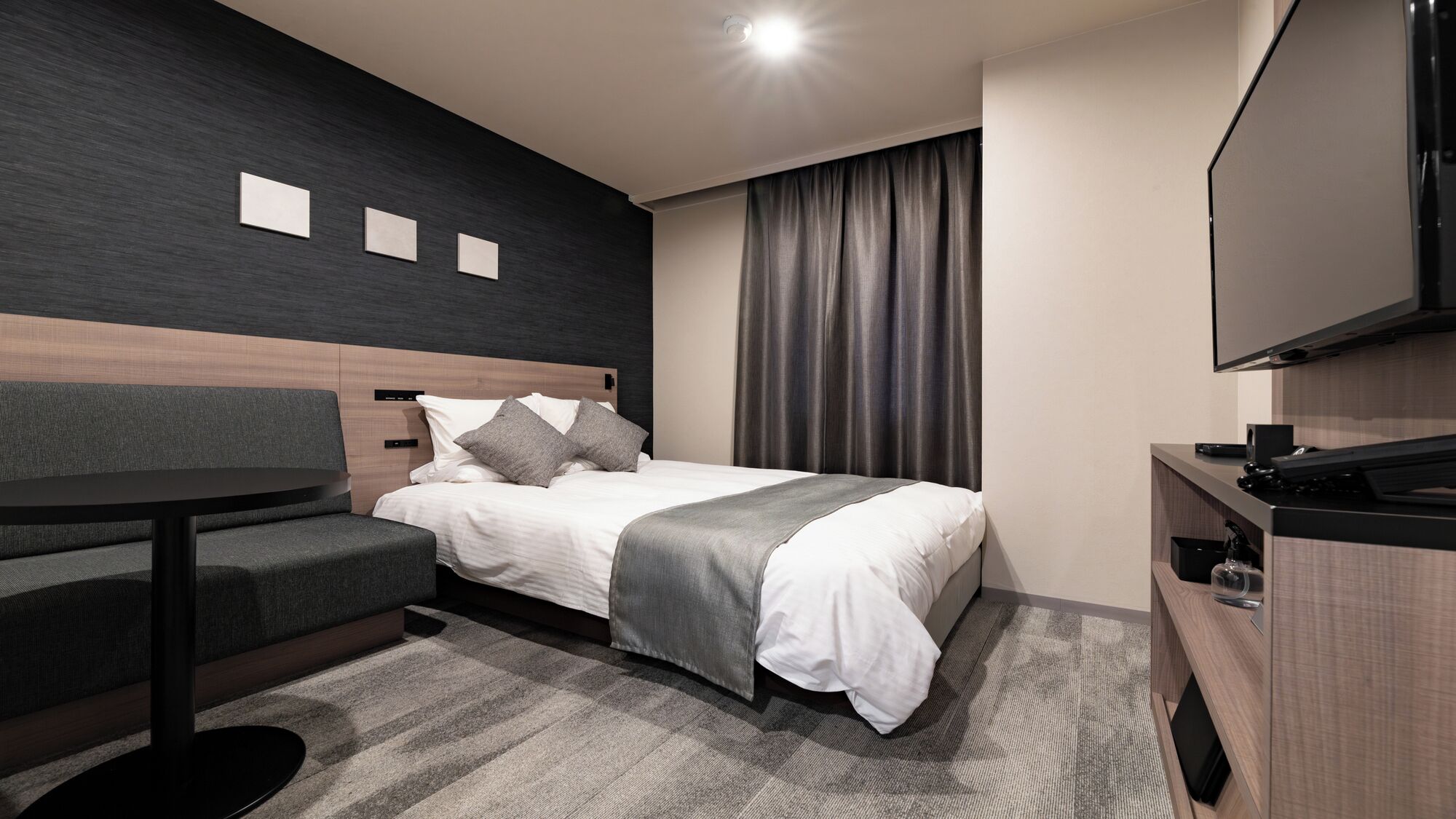 Quarto de hotel com cama, escrivaninha, cadeira, televisão e janela com cortinas.