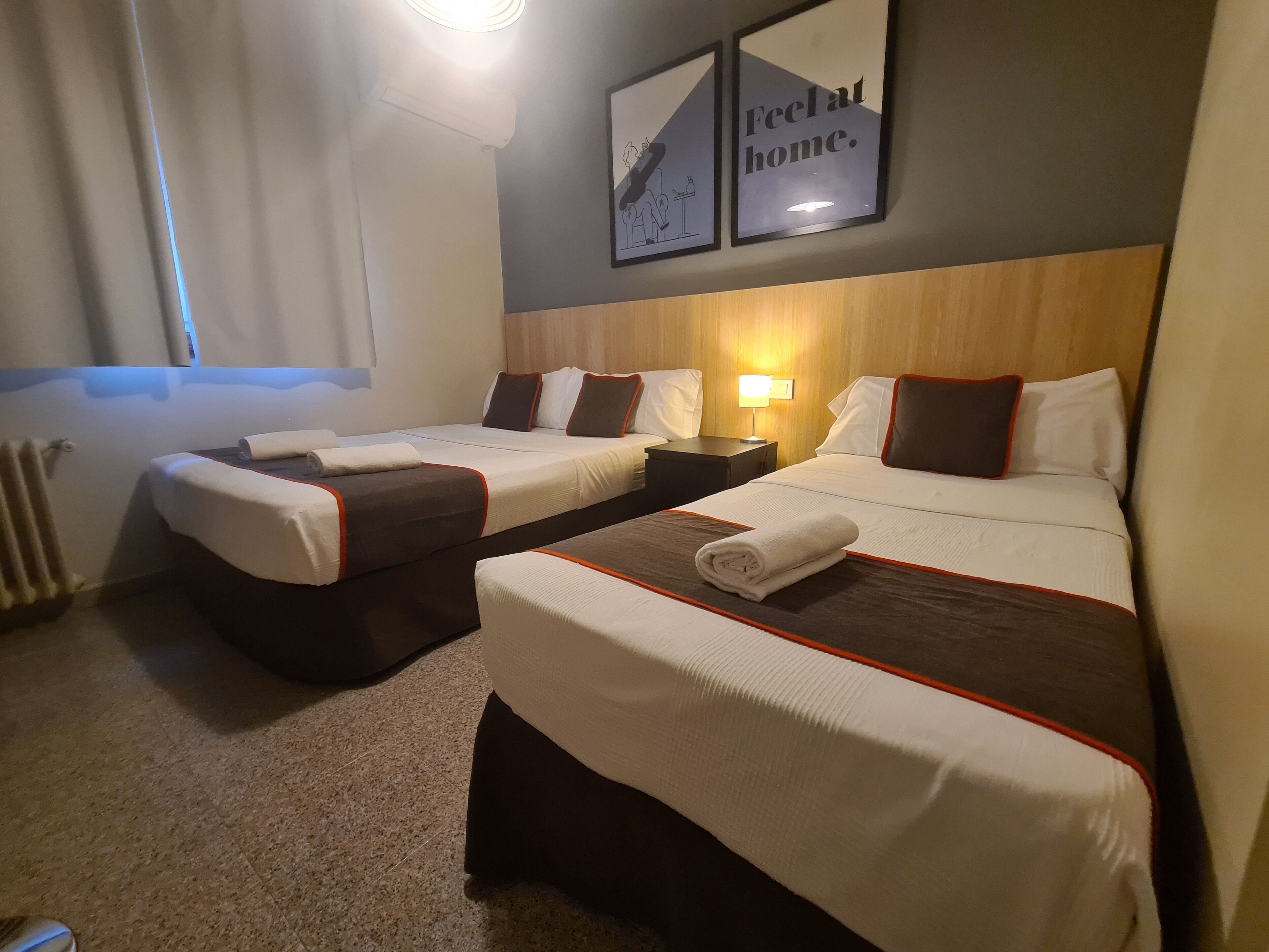 Quarto de hotel com duas camas, uma mesa de cabeceira, uma luminária e uma moldura com uma citação na parede.