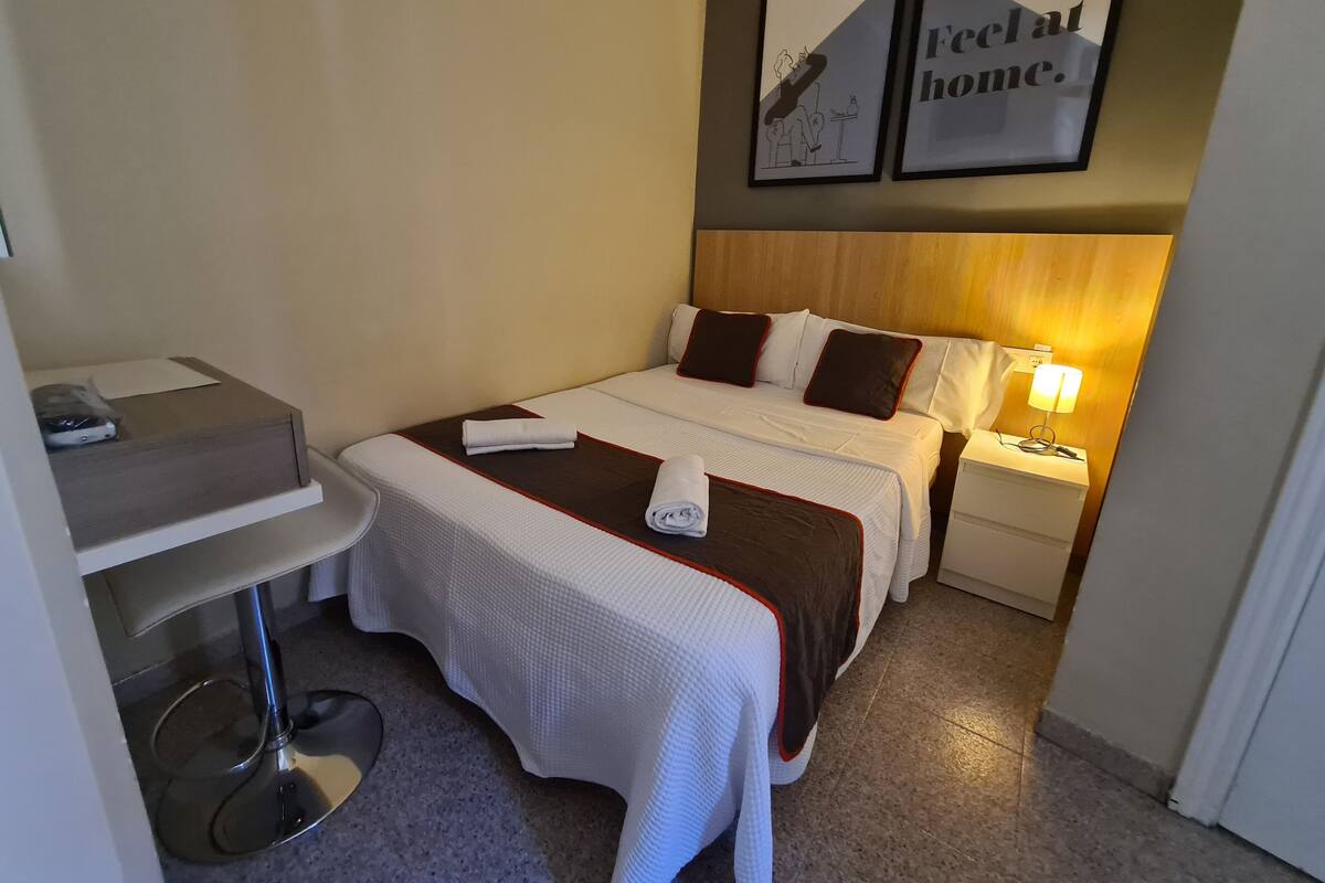 Quarto de hotel com cama, mesa de cabeceira, luminária e pequena escrivaninha com cadeira.