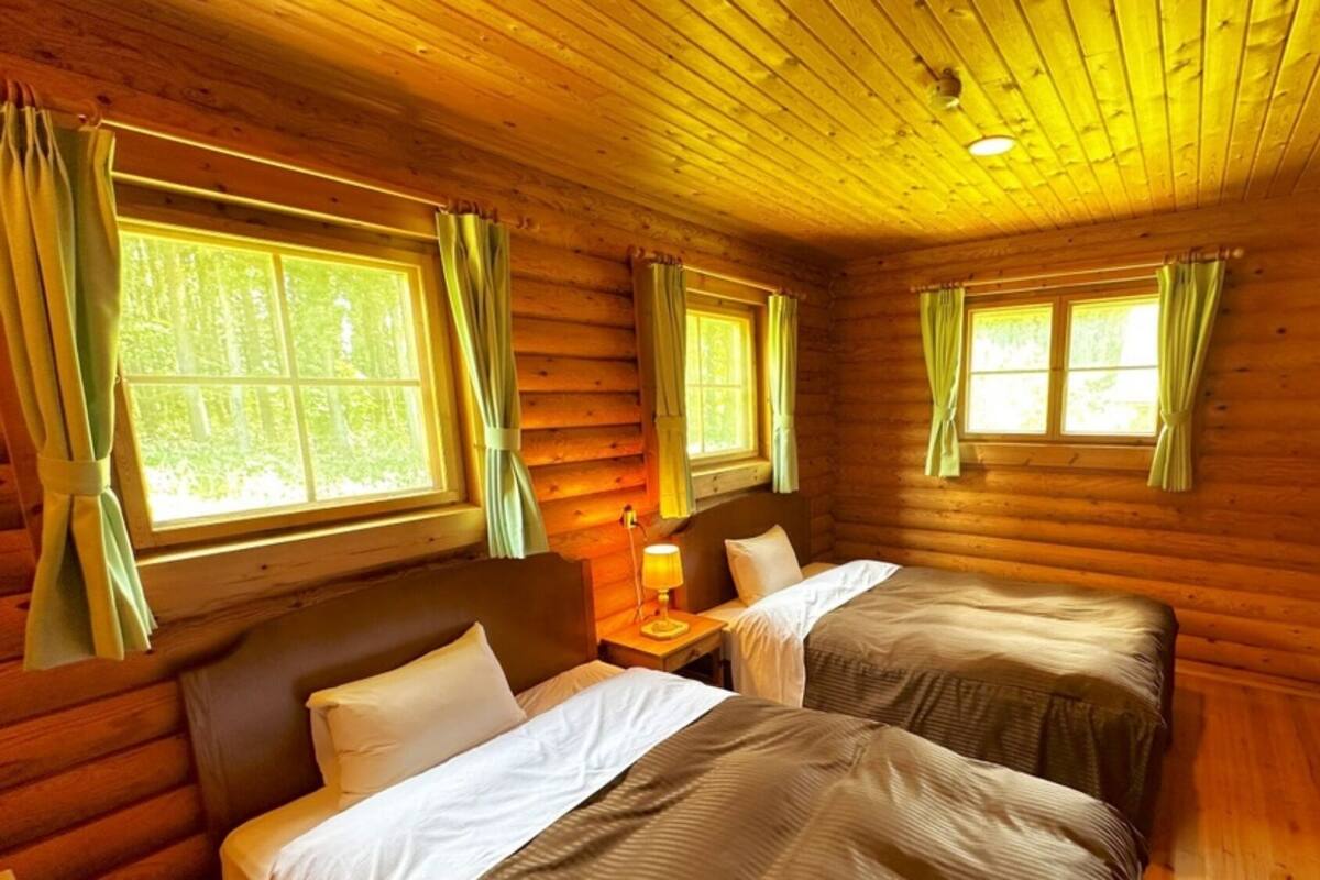 Quarto de cabana de madeira com duas camas, paredes de madeira e janelas amplas com cortinas.