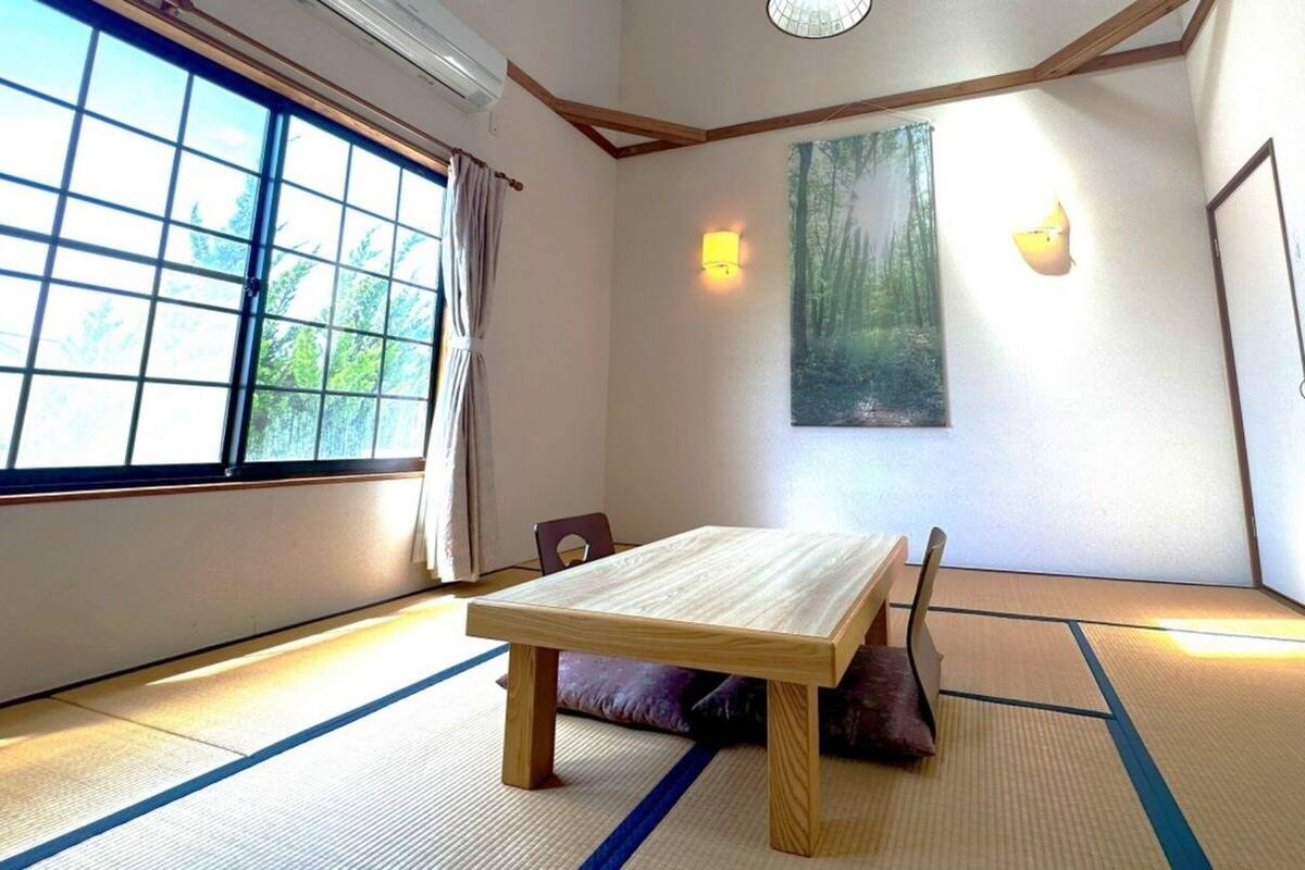 Um espaço com tatames, uma mesa de madeira baixa, tatames, uma janela com vista para árvores e duas luminárias fixadas na parede.