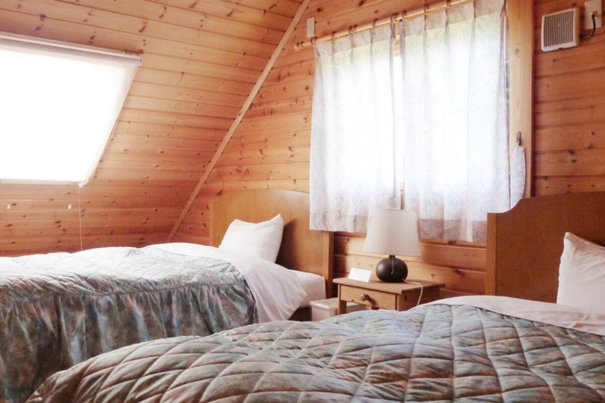 Quarto de cabana de madeira com duas camas, janela com cortinas e mesa de cabeceira com abajur.