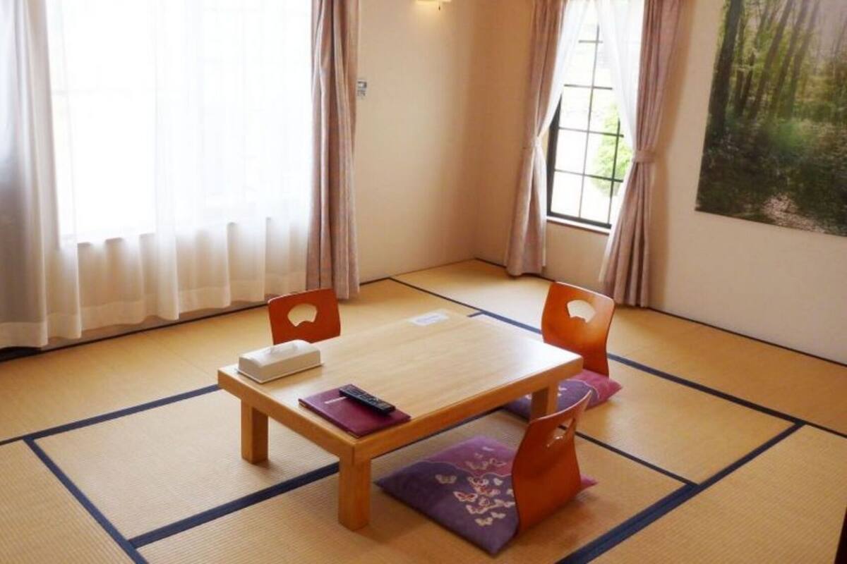 Quarto com tatame, uma mesa de madeira, duas cadeiras, uma cama e uma janela com cortinas.