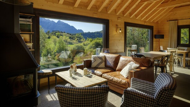 Interior acolhedor de uma cabana com uma grande janela que oferece vista para as montanhas, uma lareira e um espaço de jantar.