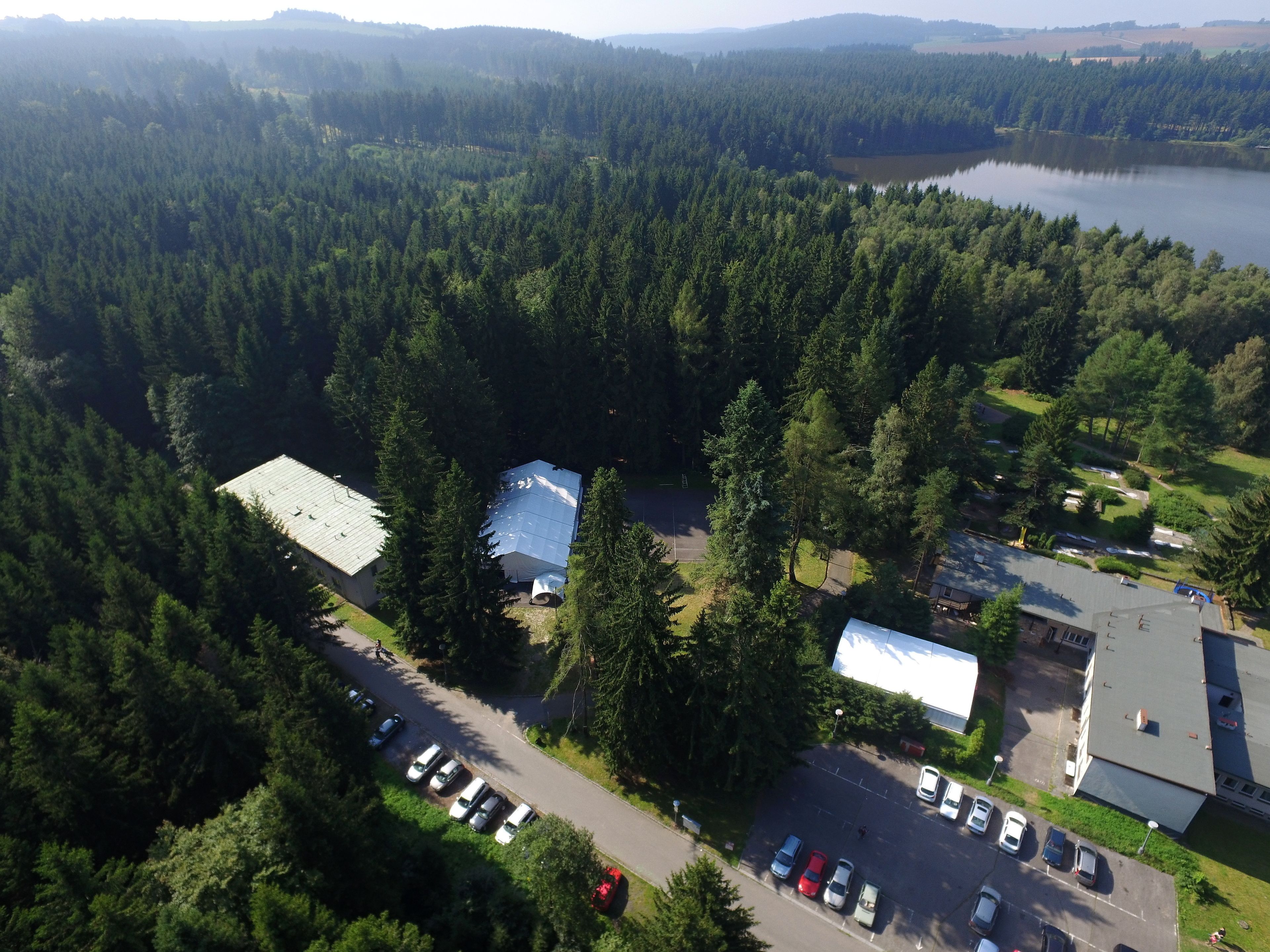 Vista aérea de uma área arborizada com um lago, um estacionamento e edifícios com telhados planos.