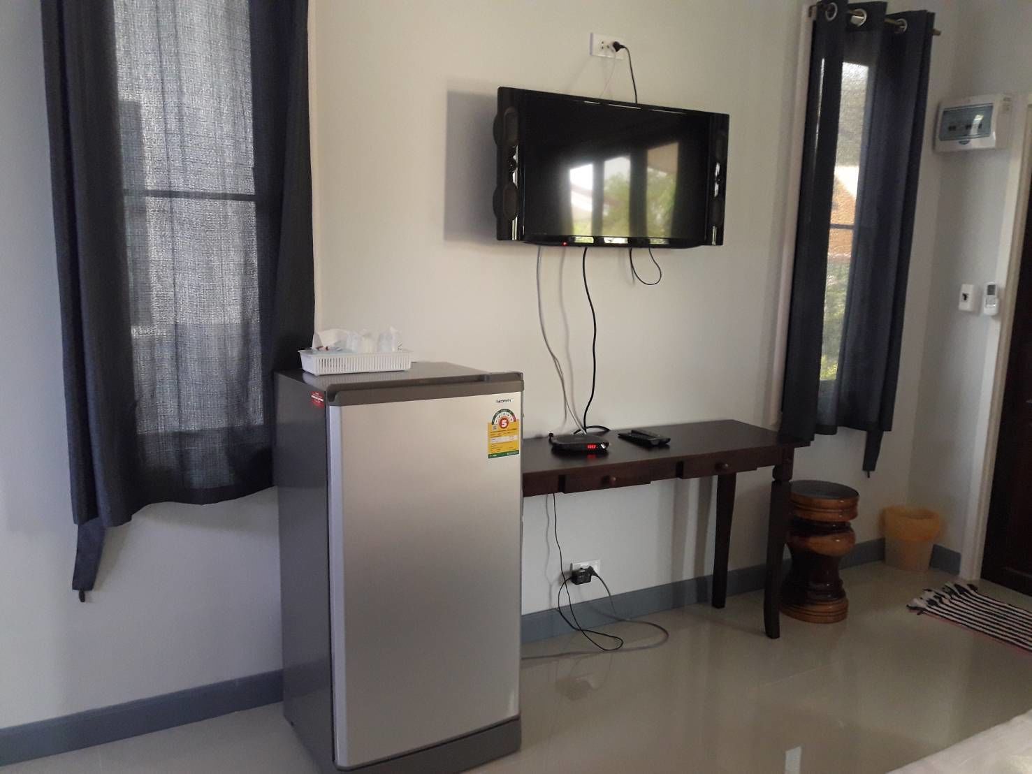 Quarto compacto com geladeira, mesa pequena, televisão fixada na parede e janela com cortinas.