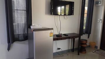 Quarto compacto com geladeira, mesa pequena, televisão fixada na parede e janela com cortinas.