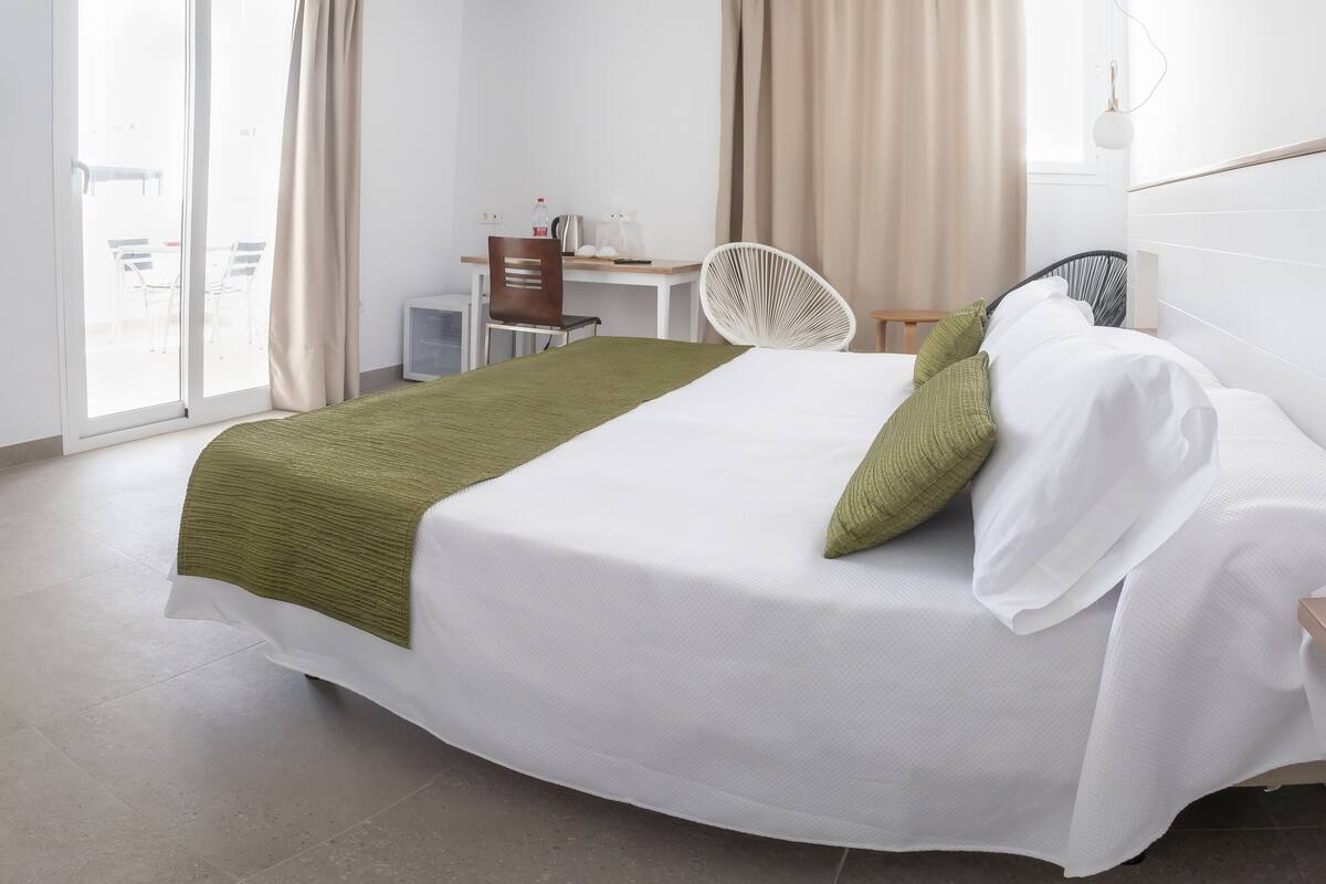 Cama bem arrumada com um manta verde, um travesseiro branco e cabeceira branca em um quarto claro com uma escrivaninha e cadeira ao fundo.