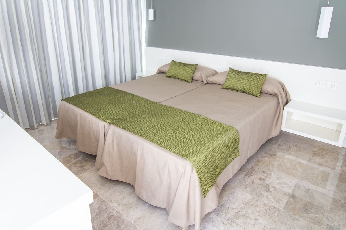 Cama bem arrumada com colcha bege e almofadas verdes, mesa de cabeceira branca e cortina na janela.