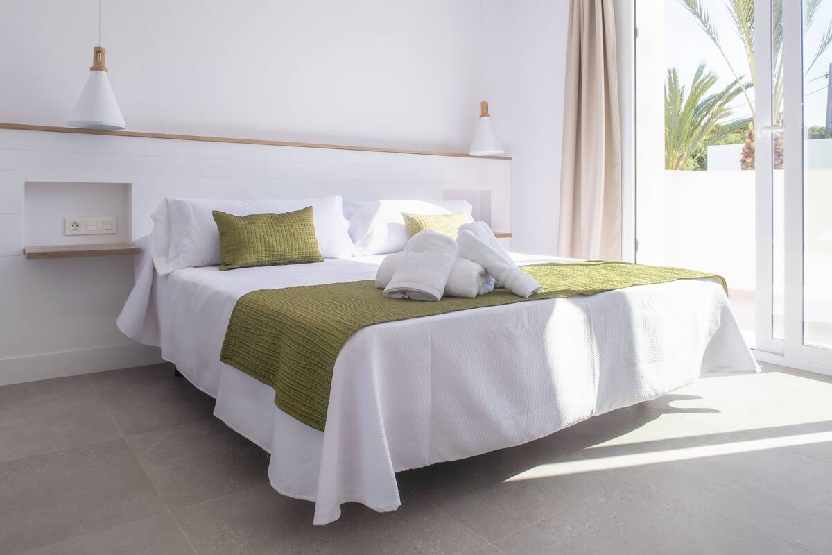 Quarto de hotel moderno com uma cama bem arrumada, uma mesinha de cabeceira e uma grande janela com vista para palmeiras.