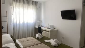 Quarto de hotel com duas camas, uma escrivaninha com computador e cafeteira, televisão na parede e janela com cortinas leves.