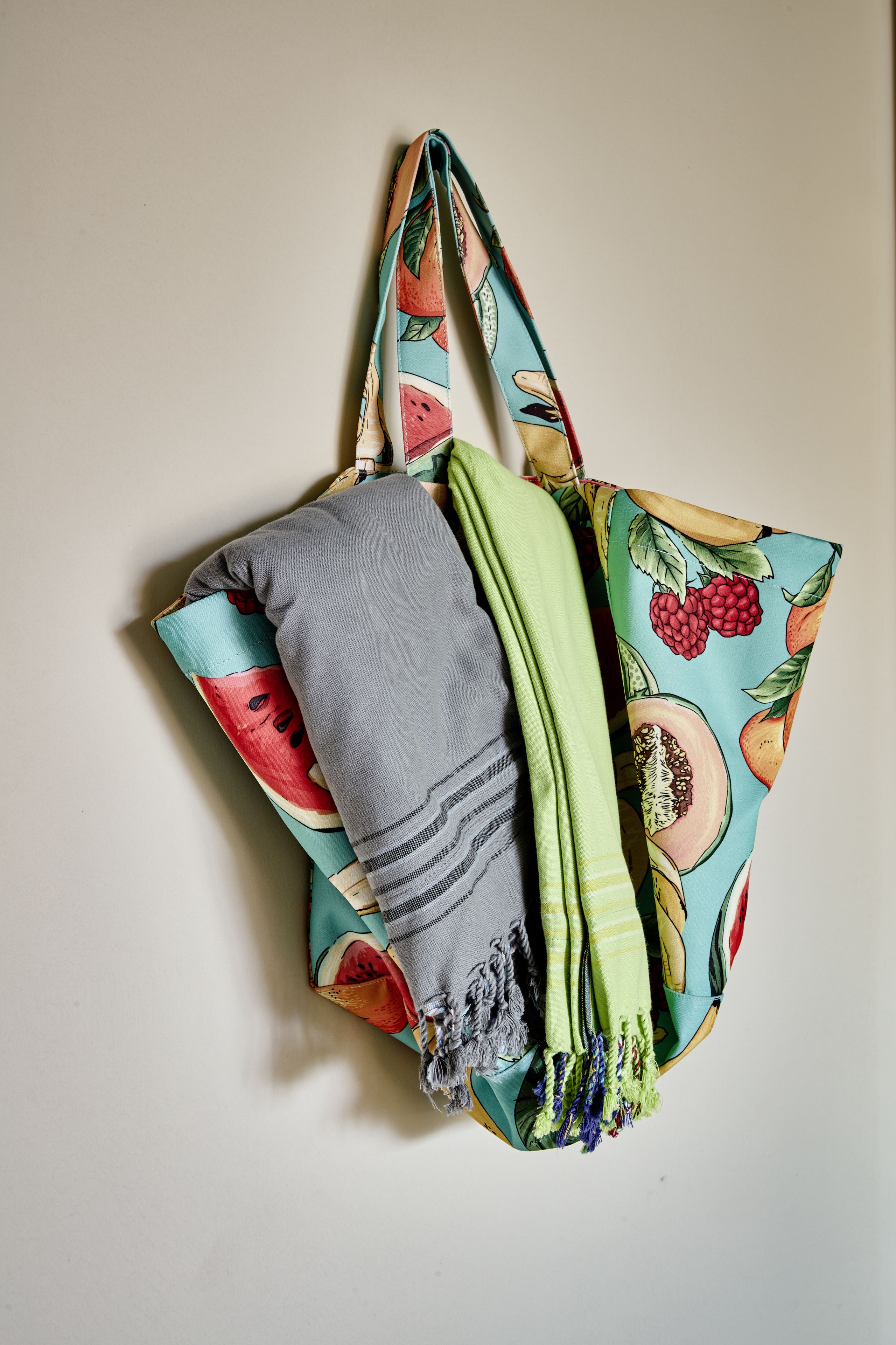 Uma bolsa colorida estampada com frutas, um travesseiro cinza e um cachecol verde.