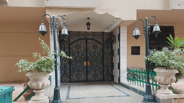 Entrada de edifício com portão decorativo, degraus de acesso e vasos de plantas de ambos os lados.