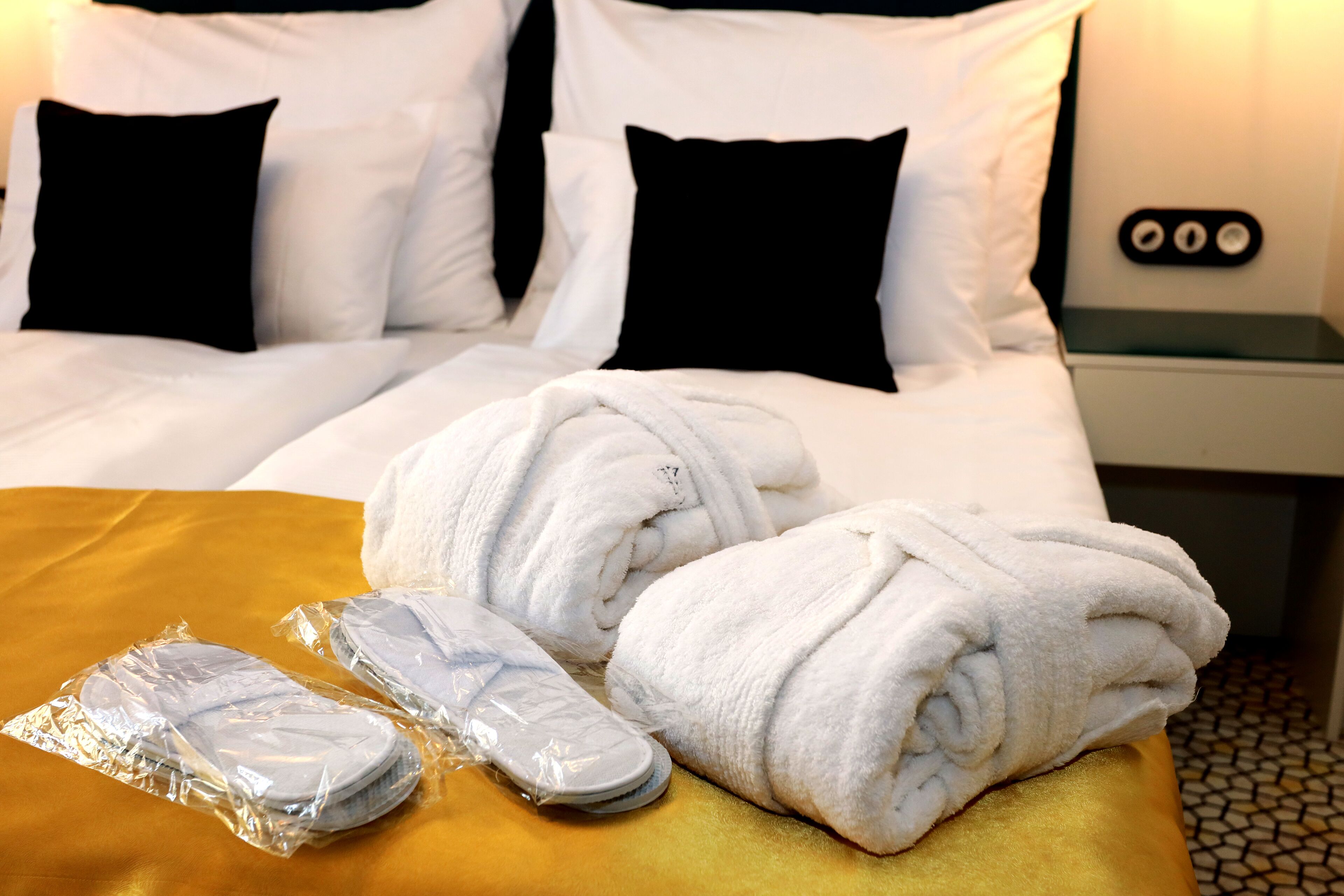 Uma cama bem arrumada com almofadas brancas e pretas, um espalhadeira dourada e dois roupões e chinelos brancos na cama.