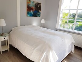 Quarto com uma cama grande, duas mesas de cabeceira com abajures, uma janela com vista para o verde e um quadro abstrato colorido na parede.