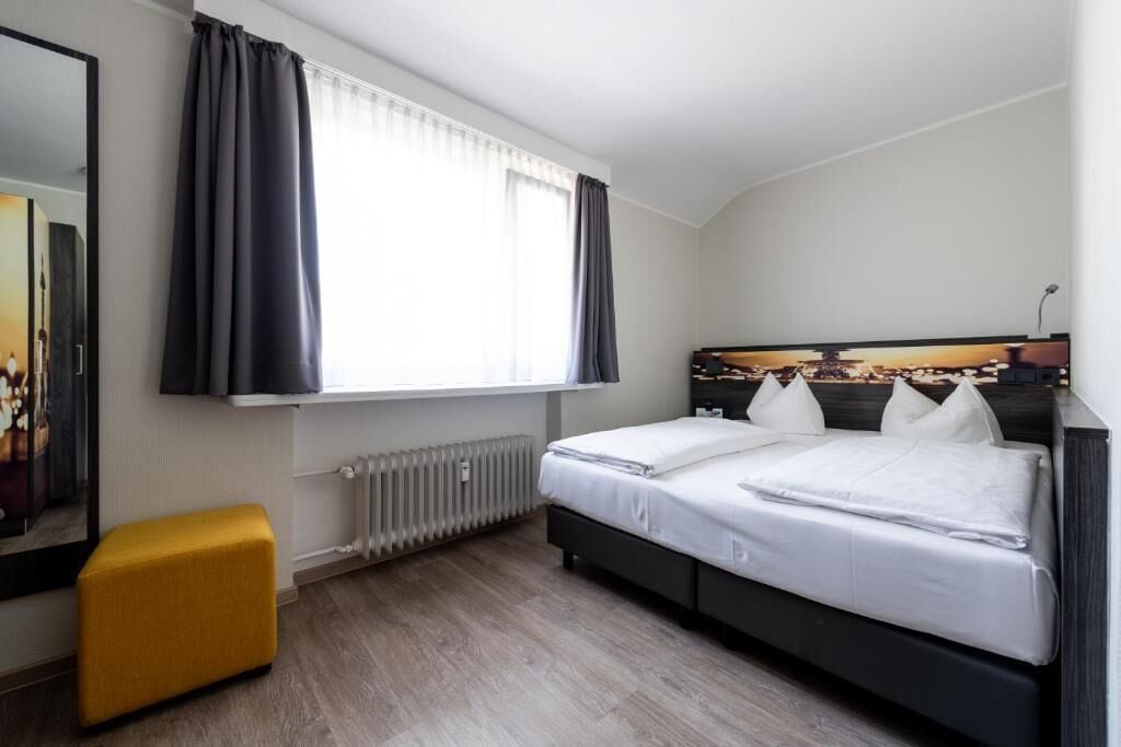 Quarto de hotel com uma cama, uma janela com cortinas, um radiador e um banquinho amarelo.