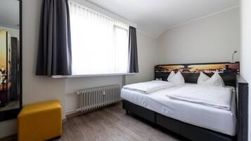 Quarto de hotel com uma cama, uma janela com cortinas, um radiador e um banquinho amarelo.