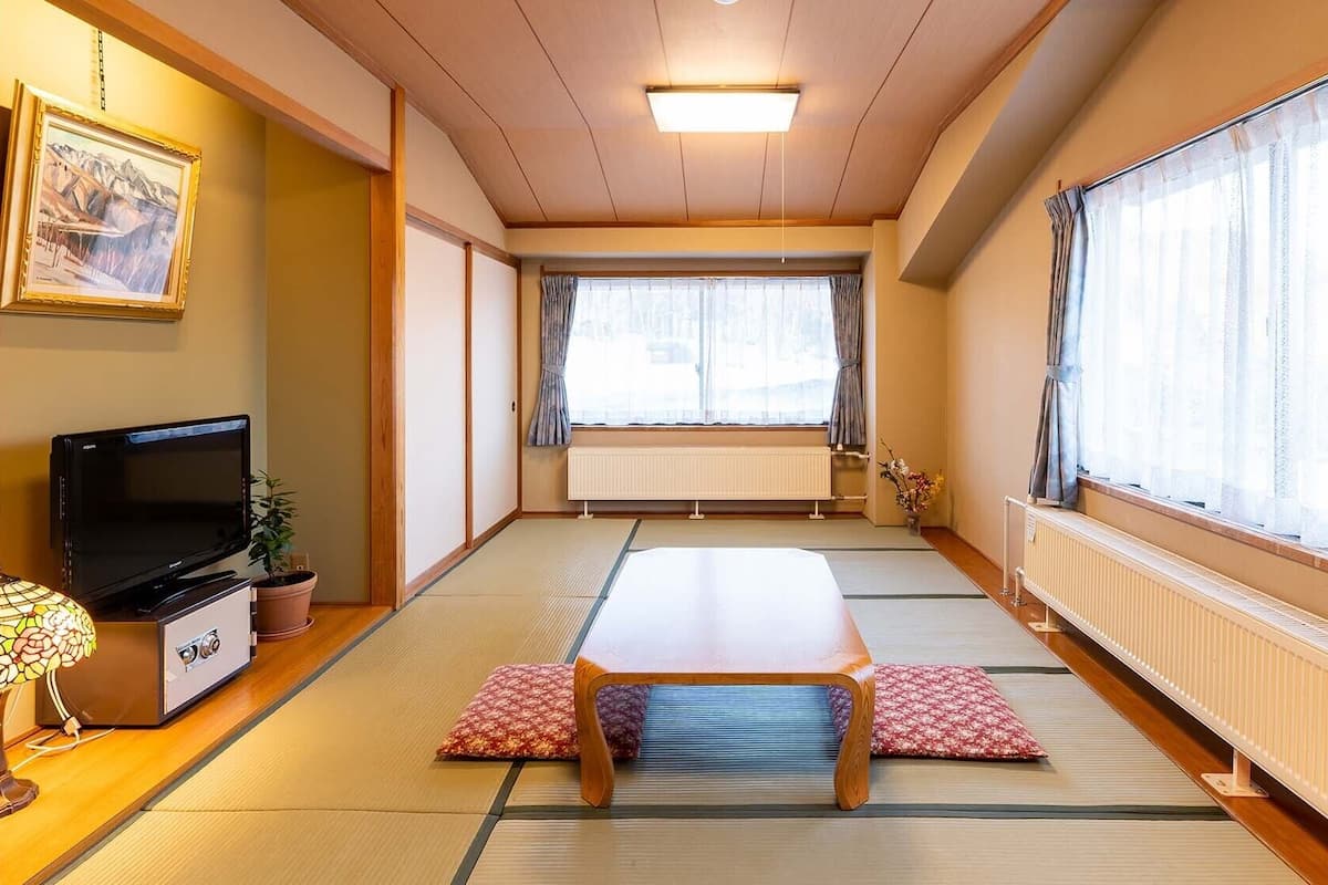 Um quarto japonês tradicional com tatames, uma mesa baixa, uma televisão e uma janela grande com cortinas.
