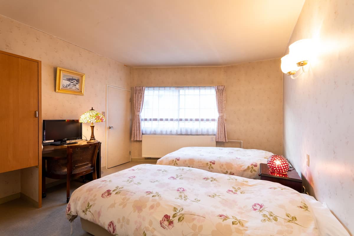 Quarto de hotel com duas camas, uma escrivaninha com luminária, televisão e janela com cortinas.