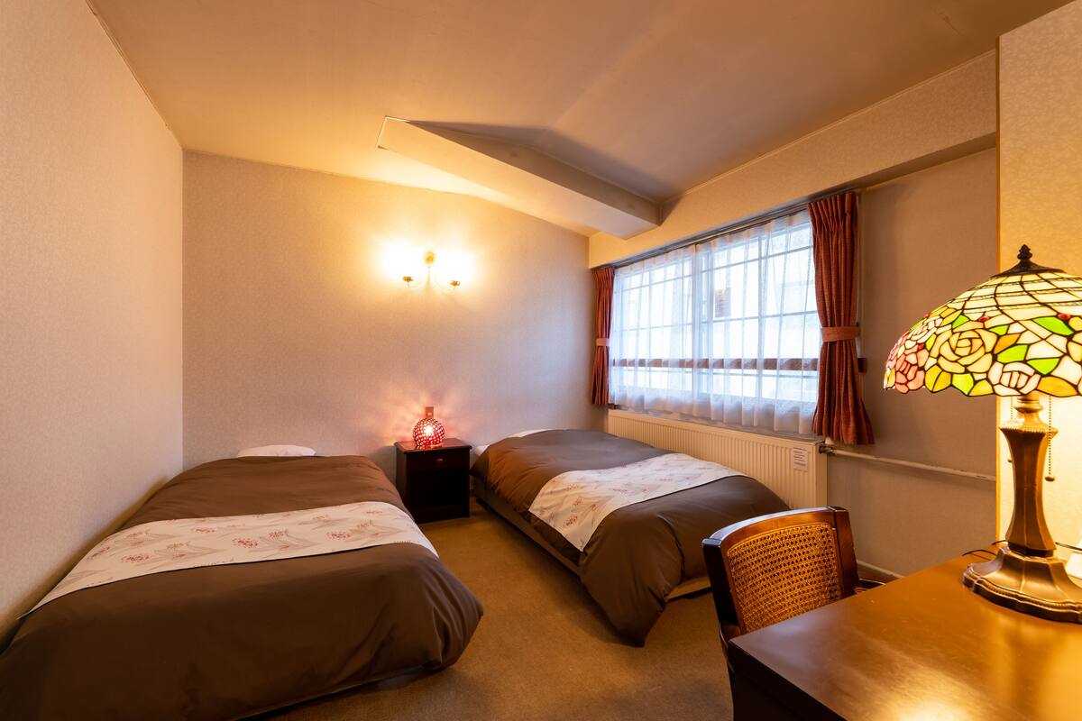 Quarto com duas camas, mesa de cabeceira com abajur, uma cadeira e janela com cortinas.