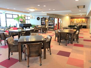 Um espaço amplo e interno com várias mesas e cadeiras, uma estante de livros e um piano.