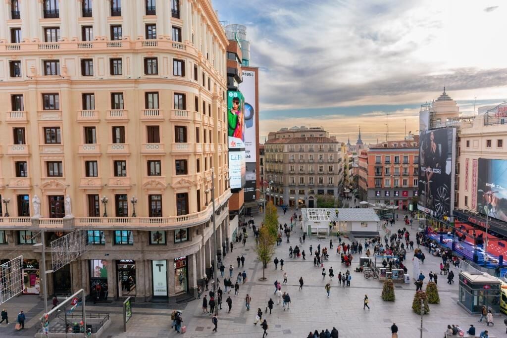 Uma praça urbana movimentada com edifícios históricos, um grande anúncio da Samsung e uma multidão de pessoas.