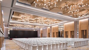 Uma sala de conferências ampla com fileiras de cadeiras brancas, uma grande televisão de tela plana e um design de teto moderno com luminárias pendentes.