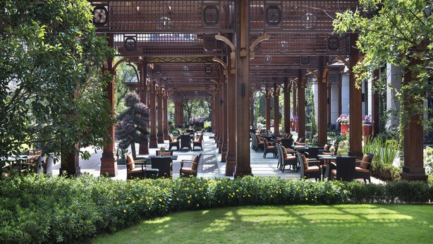 Café ao ar livre com pérgulas de madeira, mesas e cadeiras, rodeado por vegetação e flores.