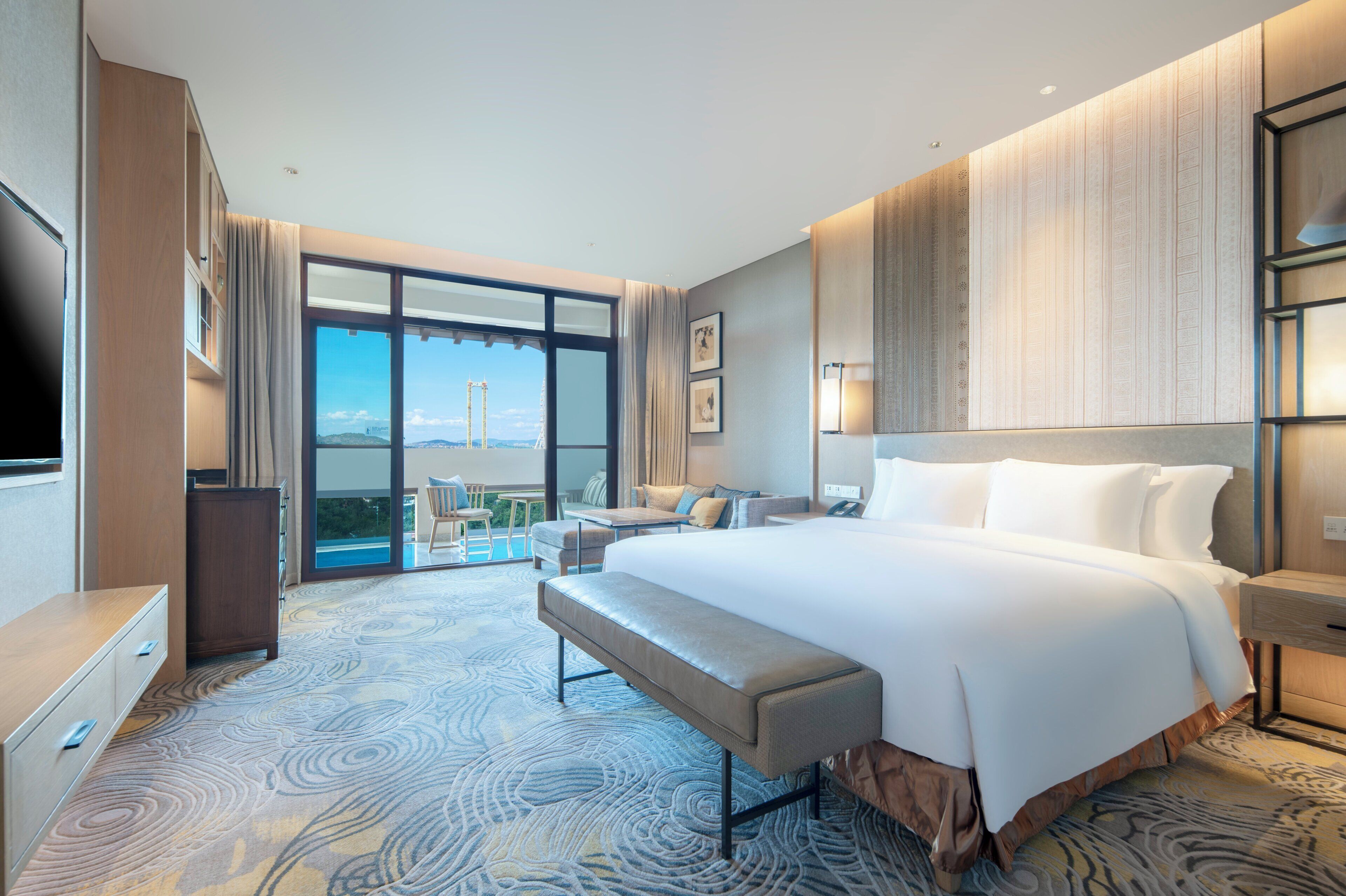 Quarto de hotel com cama grande, um banco, uma mesa e vista para a paisagem urbana através de amplas janelas.