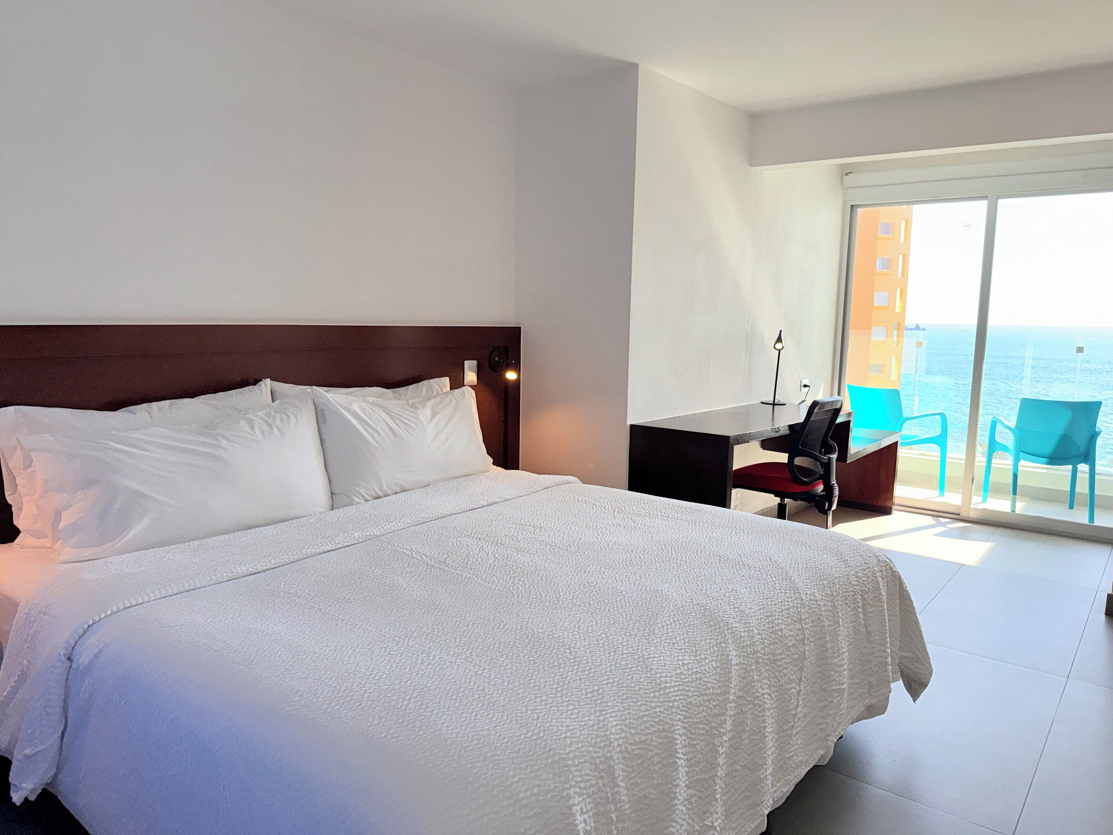 Quarto de hotel com uma cama grande, uma escrivaninha e vista para o oceano através de uma porta de correr.