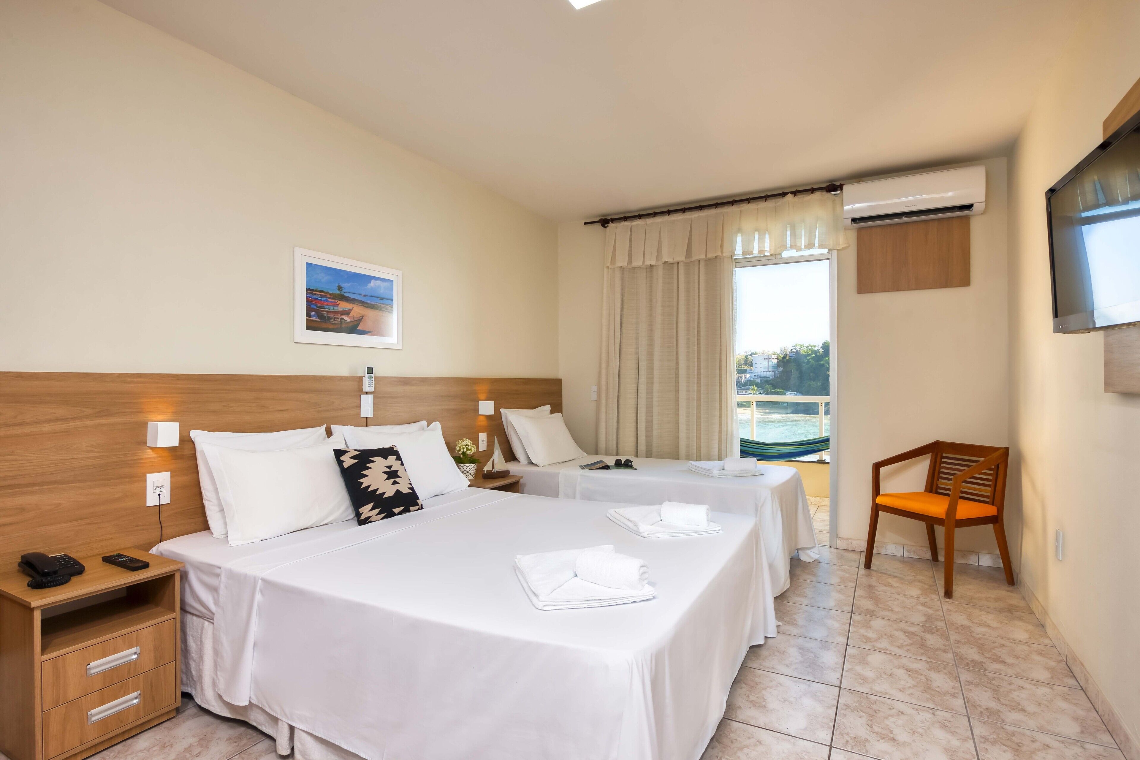 Quarto de hotel com duas camas, uma mesinha de cabeceira de madeira, uma cadeira e uma janela grande com cortinas.