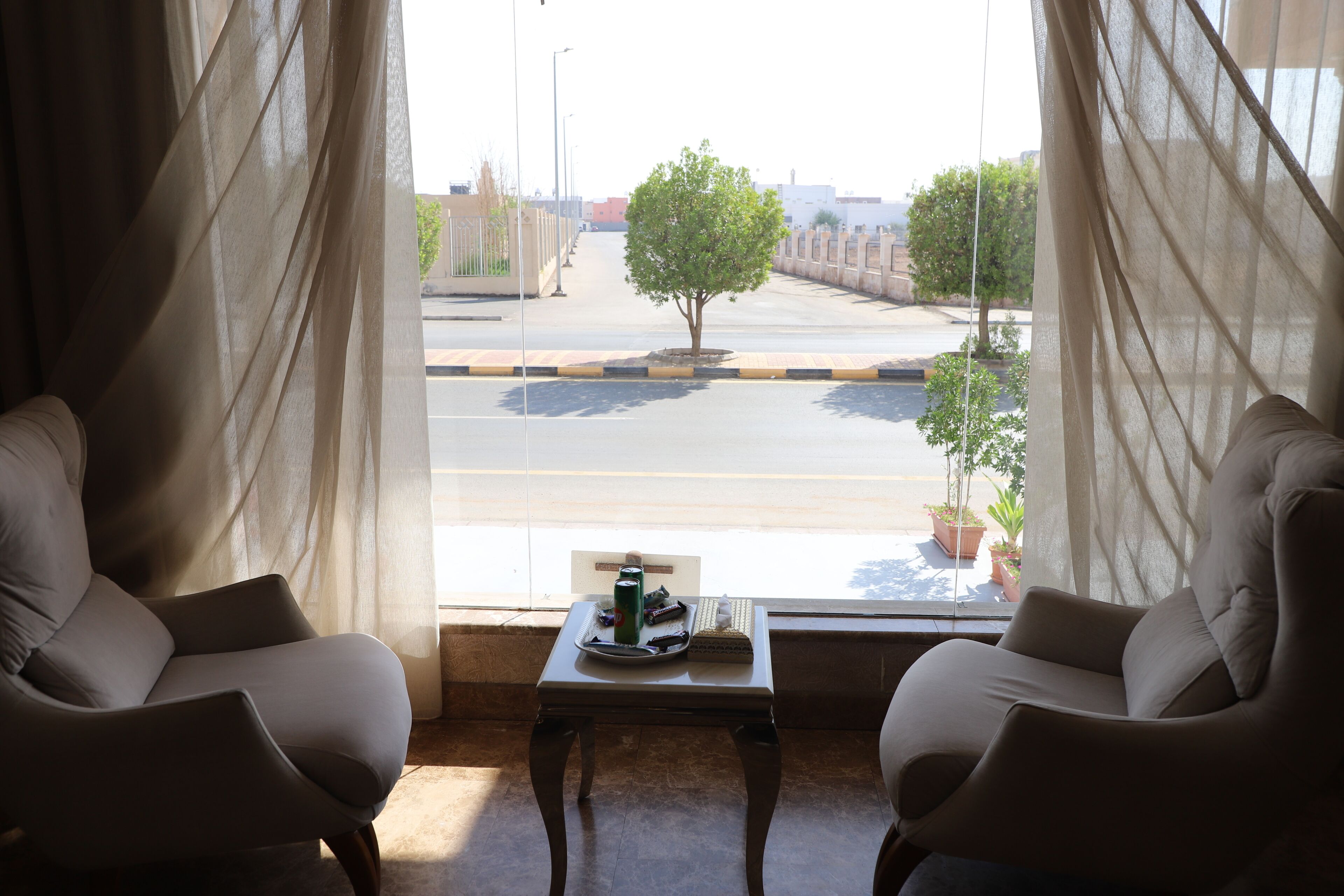 Quarto com duas cadeiras viradas para uma janela com vista para a rua. Uma pequena mesa com uma bandeja de objetos está posicionada em frente à janela.