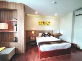 Quarto de hotel com uma cama de madeira, uma mesa de cabeceira com abajur, uma estante com objeto decorativo e uma obra de arte na parede.