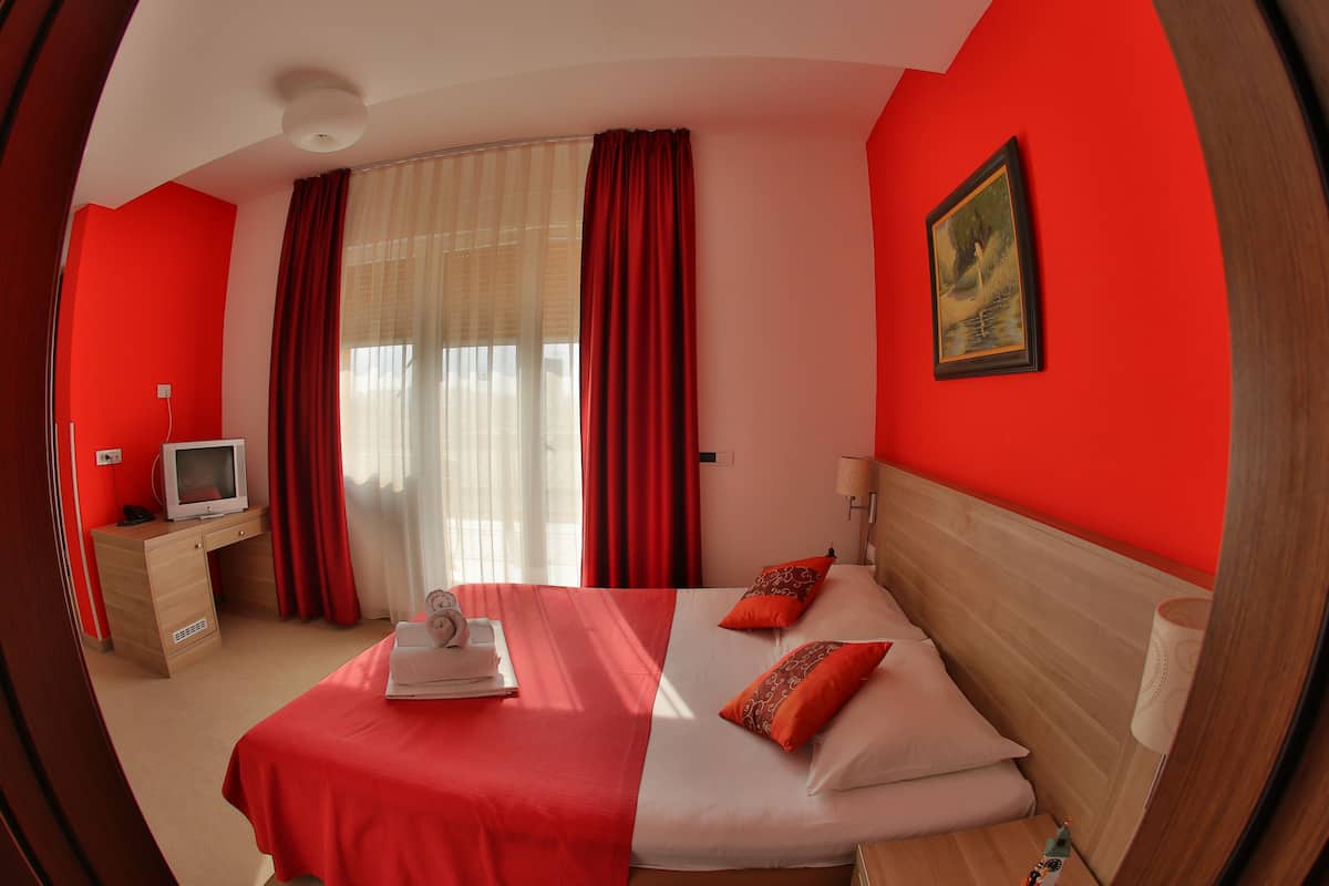 Quarto de hotel com esquema de cores vermelho e branco, cama com roupa de cama vermelha e branca, televisão em cômoda de madeira e janela com cortinas vermelhas.
