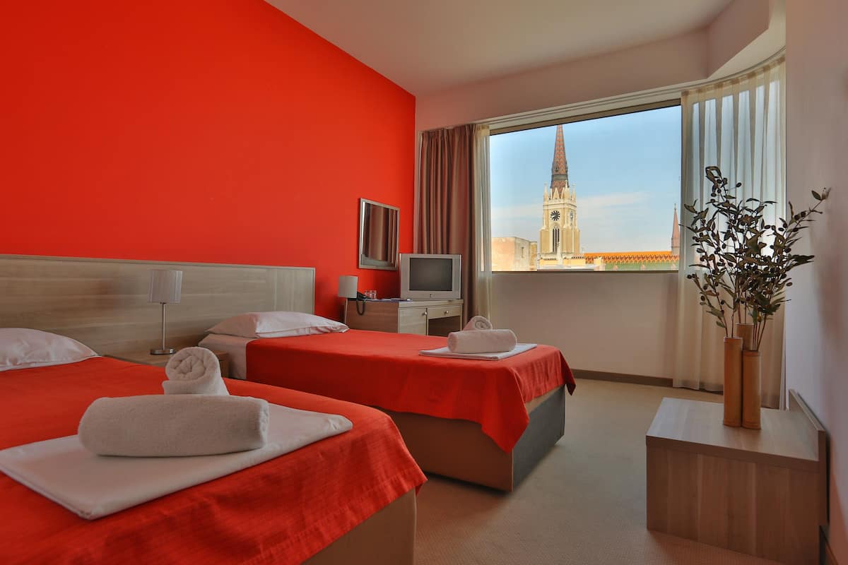 Quarto de hotel com duas camas, uma televisão, janela com vista para uma torre de igreja e uma planta em uma mesinha lateral.