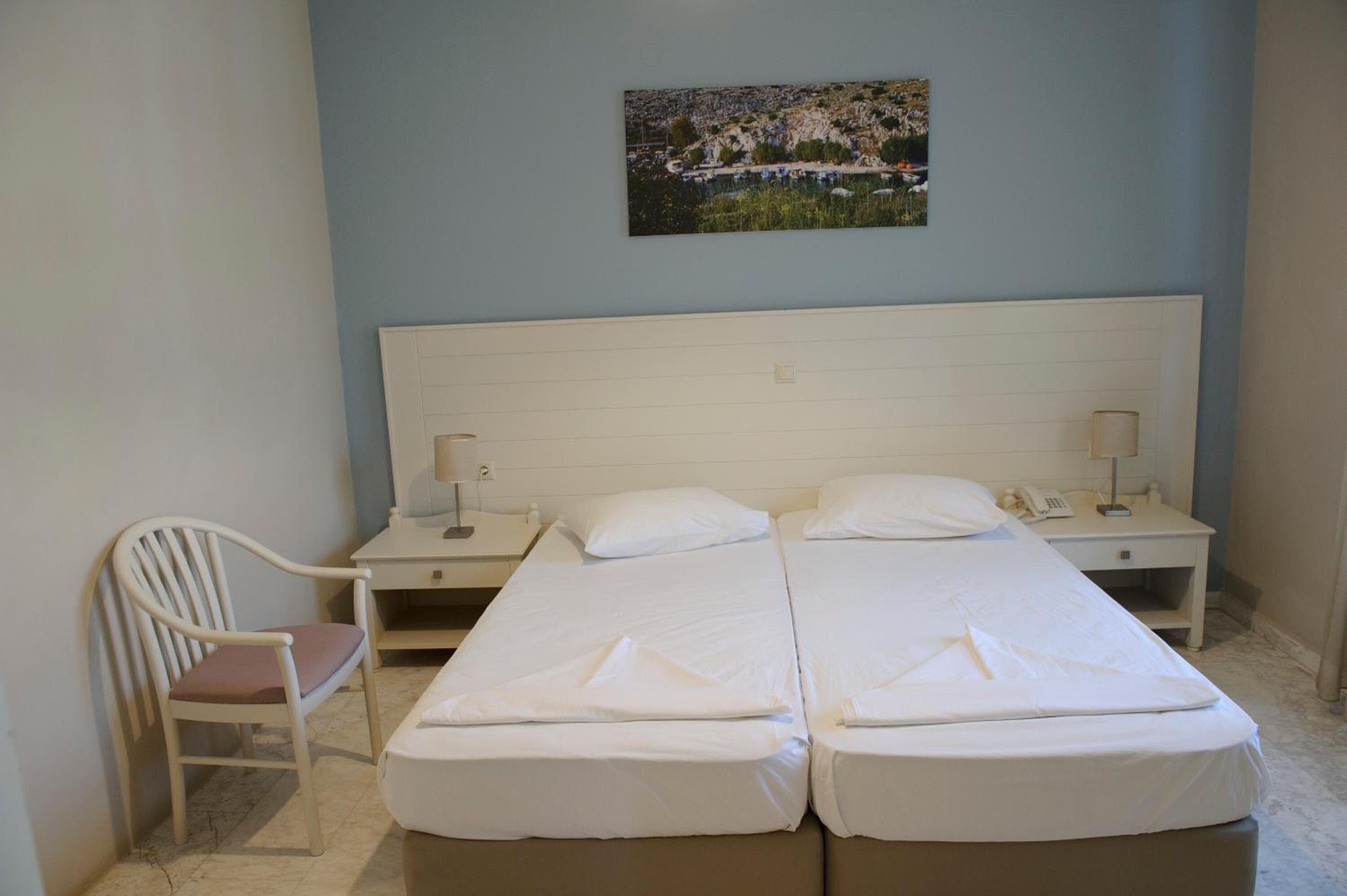 Quarto com duas camas de solteiro, uma cadeira, mesa de cabeceira com abajur e um quadro na parede.