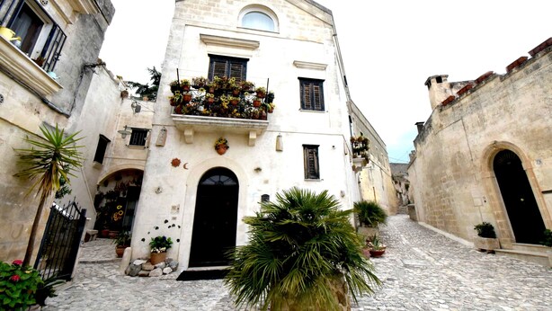 Rua de paralelepípedos estreita, ladeada por edifícios históricos com varandas decoradas com plantas em vasos.