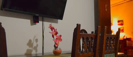 Uma sala de estar com televisão de tela plana na parede, uma mesa de jantar de madeira com cadeiras e um vaso com flores sobre a mesa.