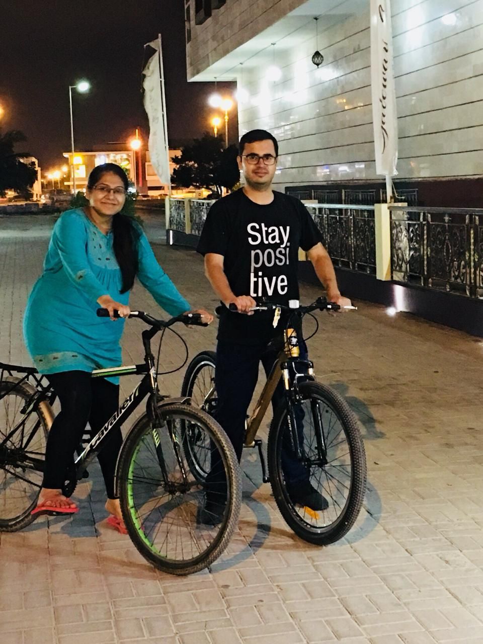 Duas pessoas de bicicleta em ambiente externo, com calçada pavimentada e um edifício ao fundo.