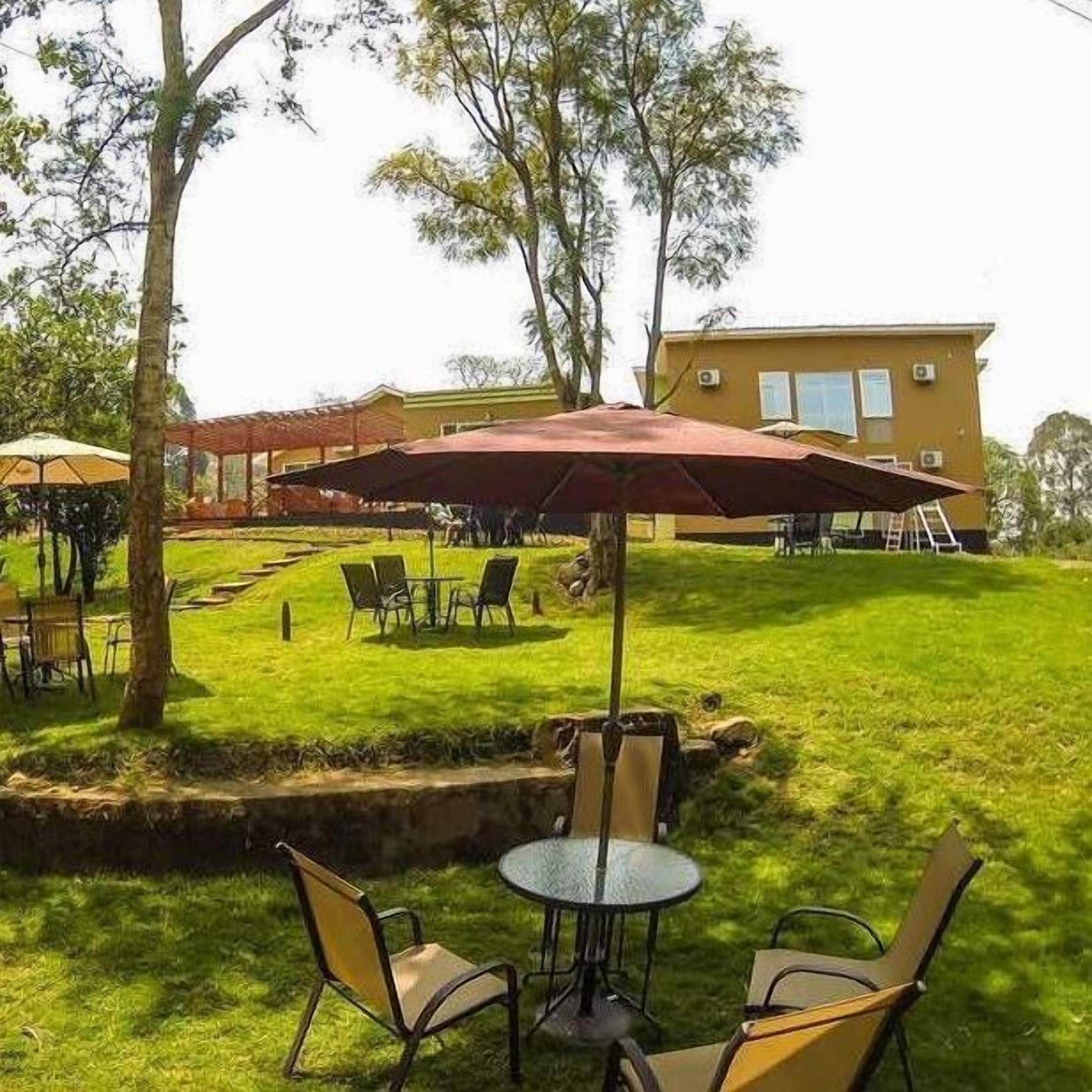 Uma área externa gramada com mesas e cadeiras, um grande guarda-sol e um edifício ao fundo.