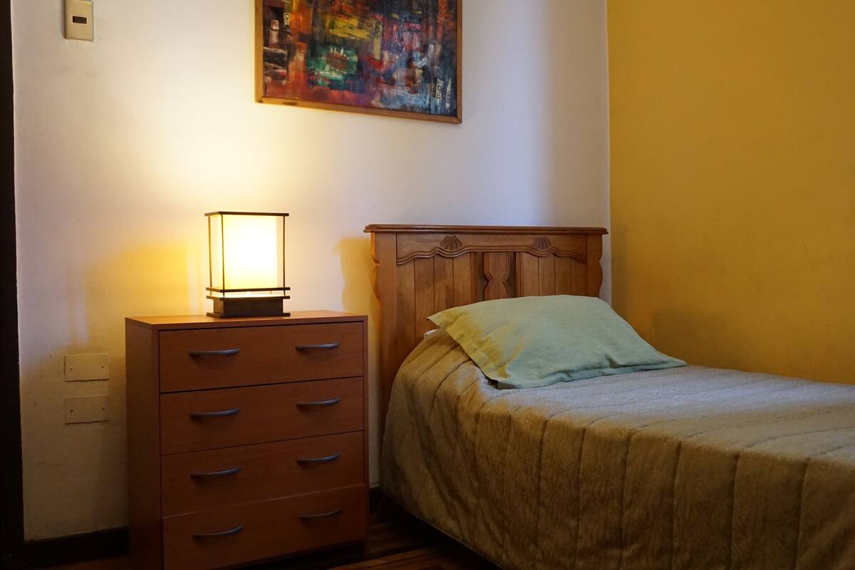 Quarto com cama, mesa de cabeceira de madeira com gavetas, luminária e um quadro na parede.